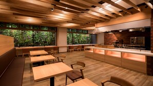 Um restaurante moderno com mesas e cadeiras de madeira, parede de tijolos e grandes janelas com vegetação.