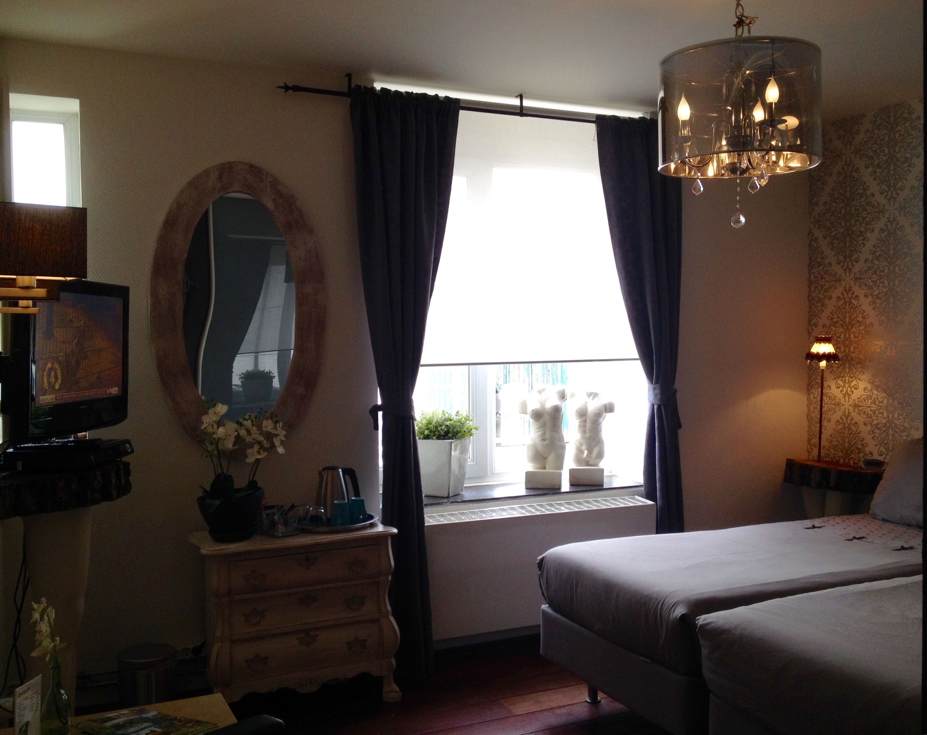 Quarto com cama, cômoda, espelho, janela com cortinas e um lustre.