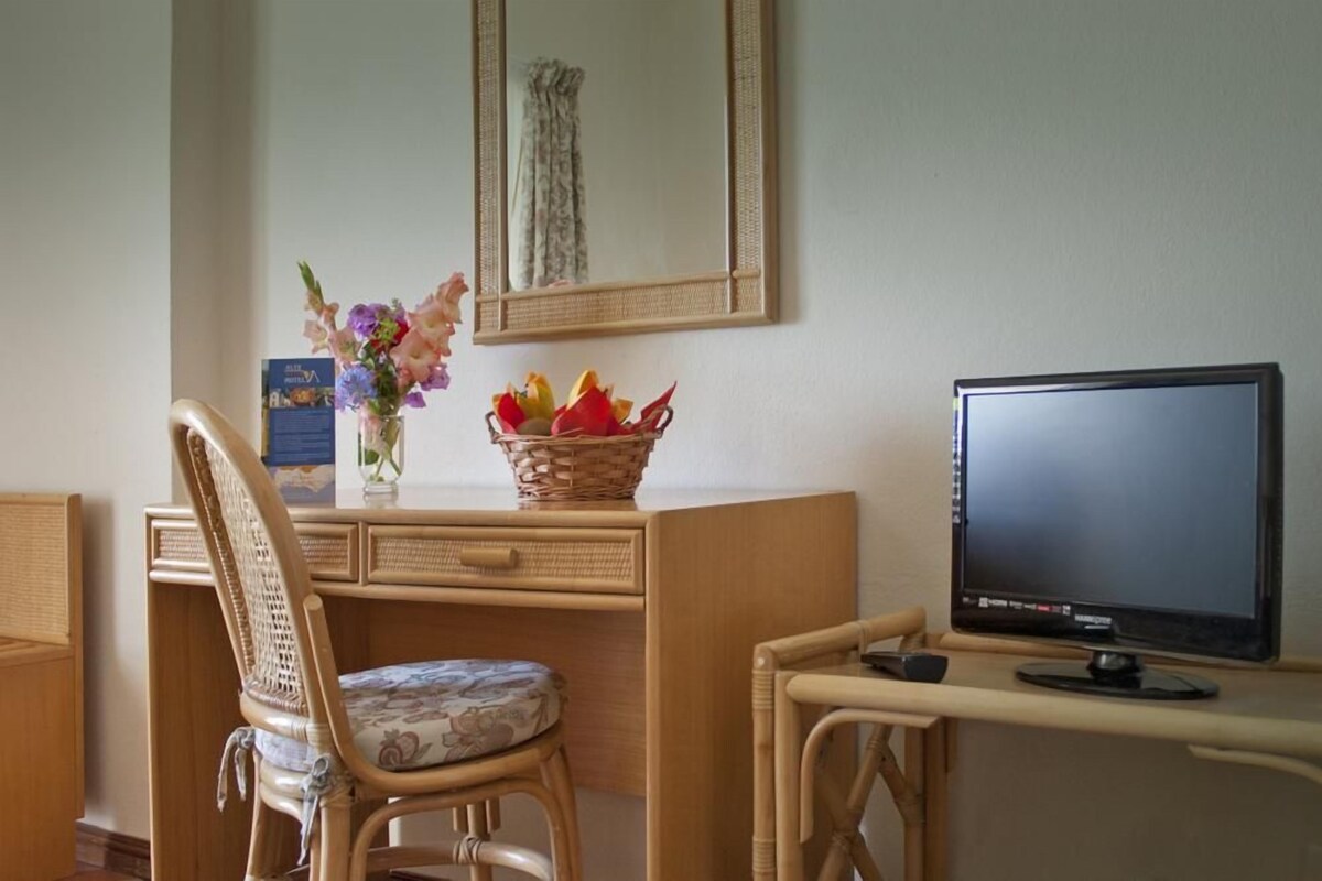 Uma escrivaninha de madeira com um monitor de computador, uma cadeira de vime, uma cesta de frutas e um vaso de flores.