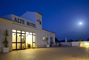 Exterior do Hotel Alte, com entrada bem iluminada, acesso por portão e céu noturno claro.