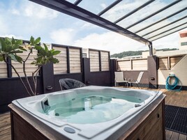 Uma banheira de hidromassagem com um deck de madeira, um espaço de varanda coberto e vista para os edifícios ao redor.