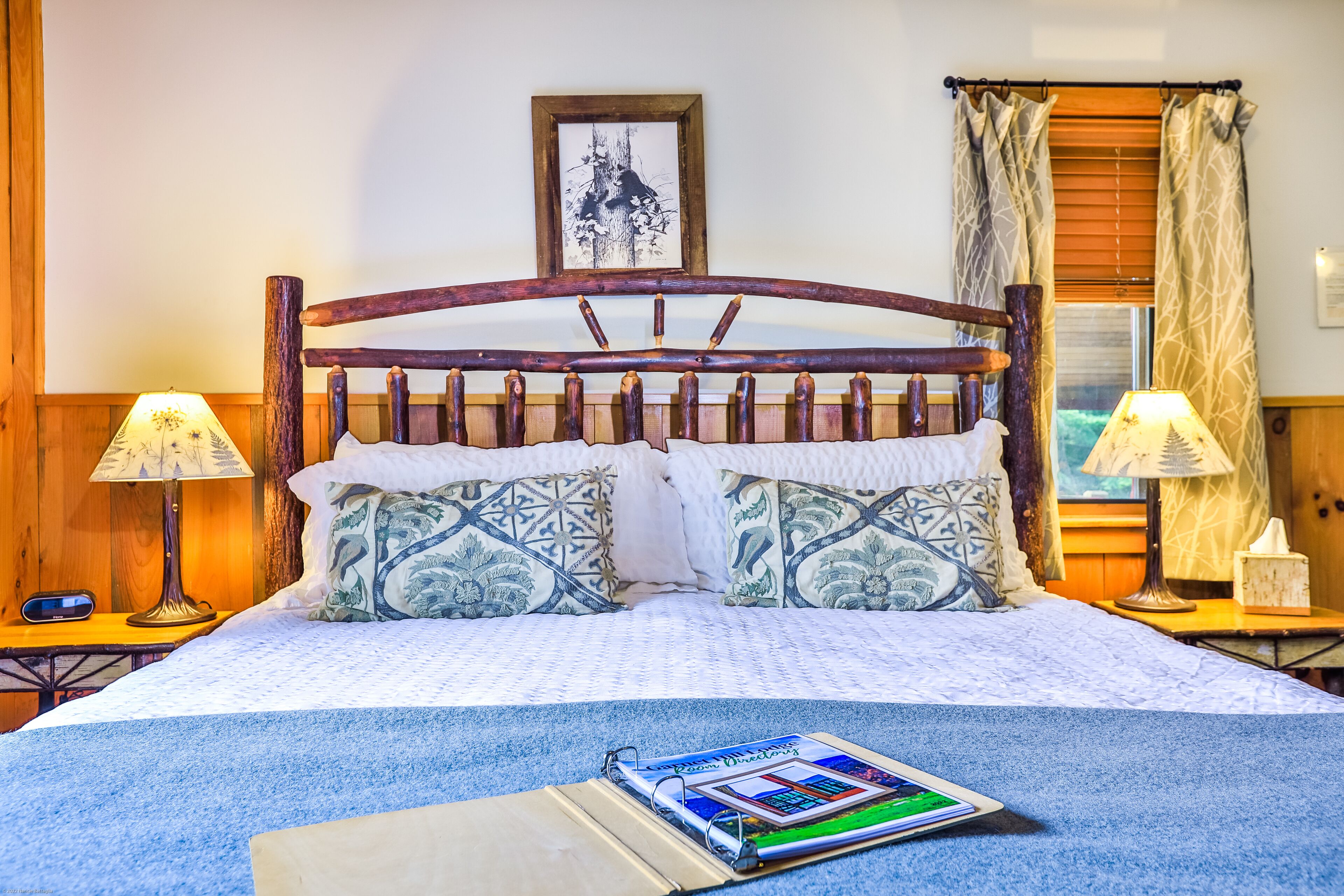 Uma cama bem arrumada com almofadas estampadas, cabeceira de madeira e mesa de cabeceira com abajur e um livro.