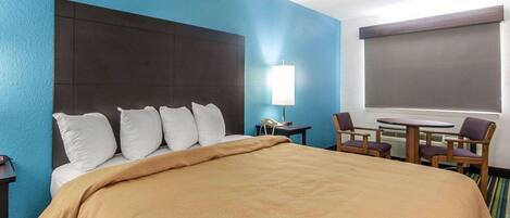 Quarto de hotel com uma cama grande, uma escrivaninha, duas cadeiras, uma janela com persianas e uma mesa de cabeceira com abajur.