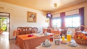 Uma sala de jantar com mesas preparadas para o café da manhã, incluindo pratos, xícaras e uma variedade de frutas e bebidas.