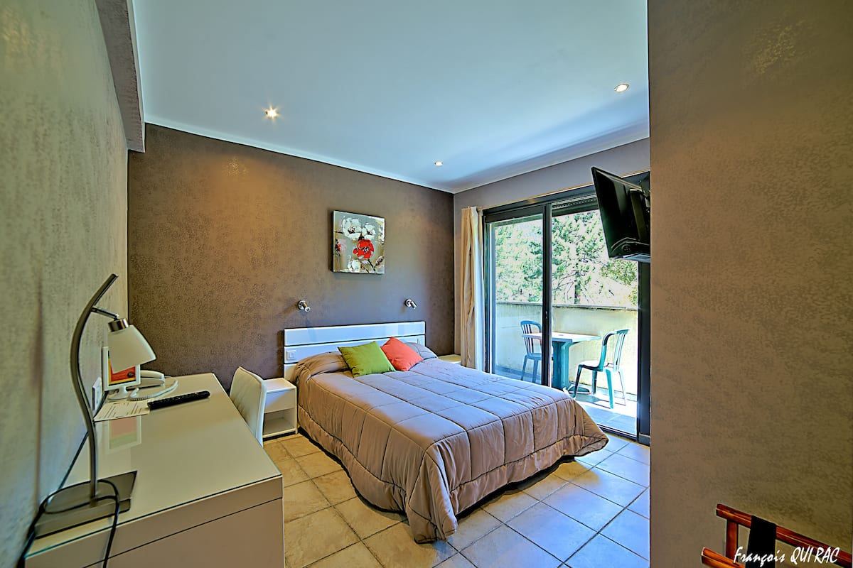 Quarto com cama, escrivaninha, cadeira e televisão fixada na parede.