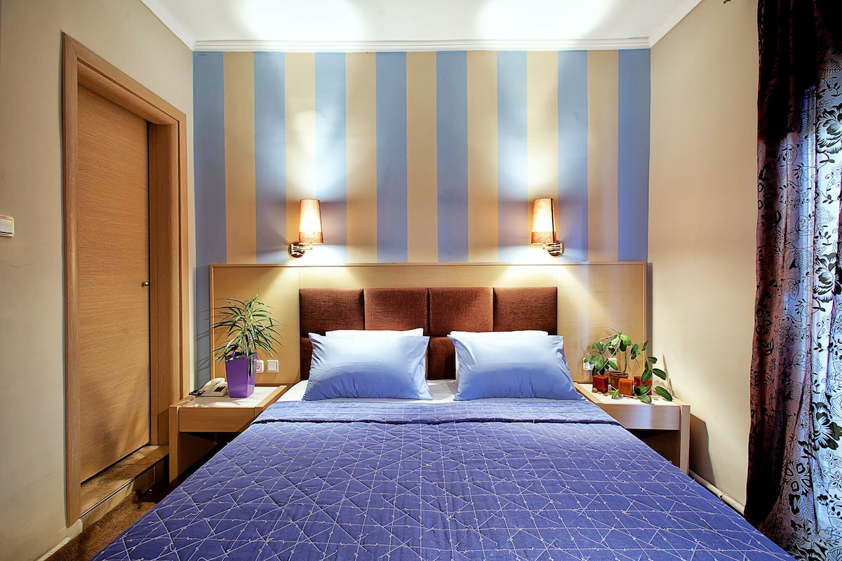 Quarto de hotel com uma cama grande, duas mesas de cabeceira, um criado-mudo, uma luminária na parede e uma porta.