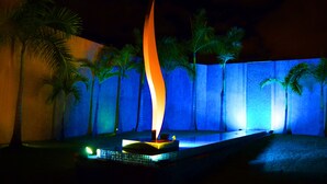 Escultura ao ar livre noturna com uma estrutura alta e iluminada no centro, rodeada por palmeiras e uma parede de azulejos.