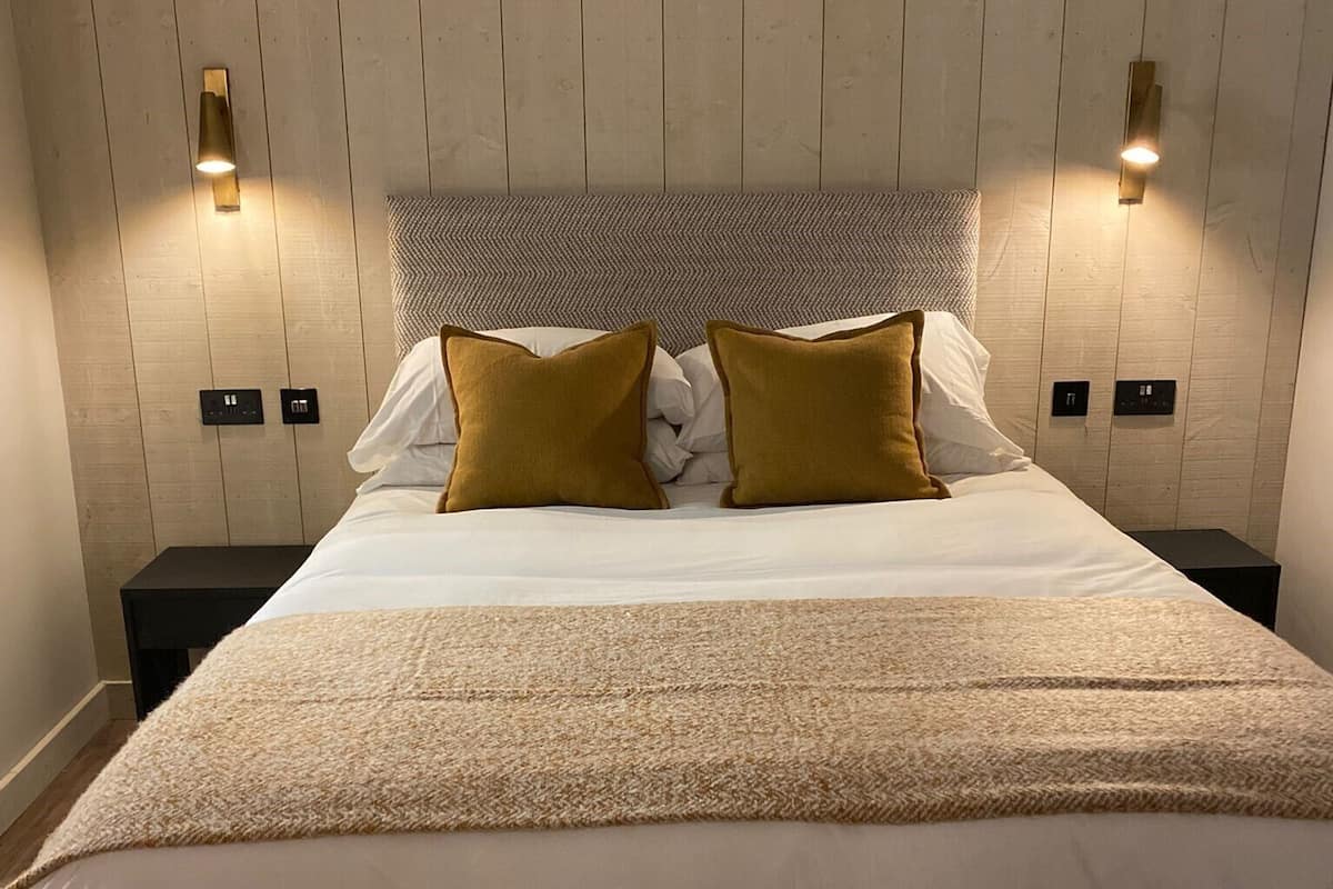 Quarto moderno com cabeceira de madeira, duas mesas de cabeceira com abajures fixados na parede, uma cama grande com manta bege e duas almofadas.