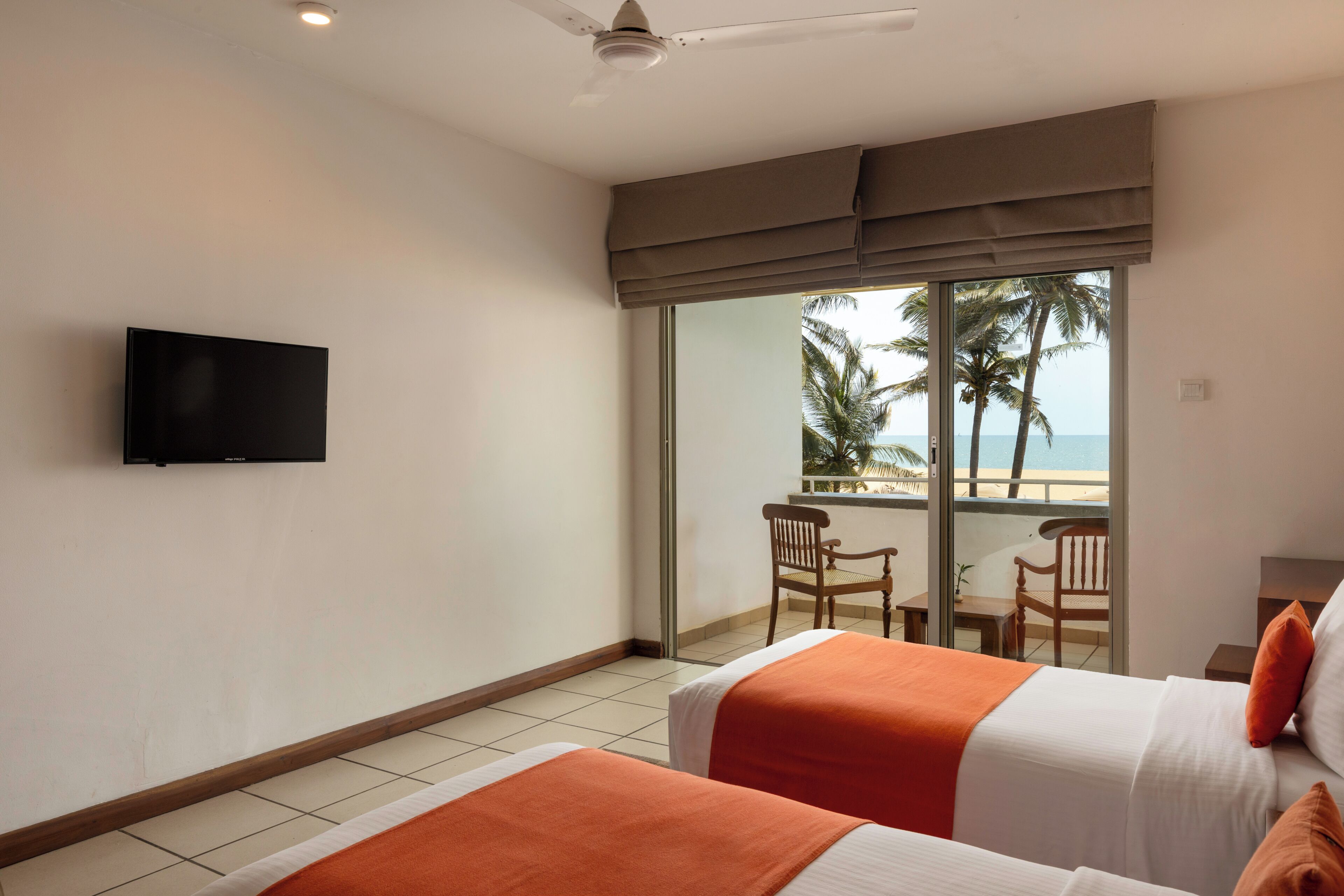 Quarto de hotel com duas camas, televisão, varanda com mesa e cadeiras, e vista para a praia.