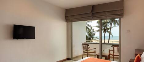 Quarto de hotel com duas camas, televisão, varanda com mesa e cadeiras, e vista para a praia.