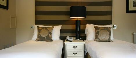 Quarto de hotel com duas camas, cabeceira com listras horizontais, luminária de cabeceira e uma pequena mesa de cabeceira branca com gavetas.