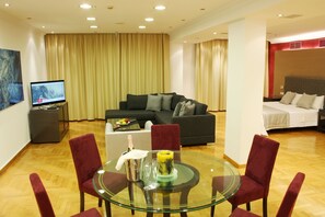 Quarto de hotel moderno com mesa de jantar de vidro, cadeiras vermelhas, sofá, cama e televisão.