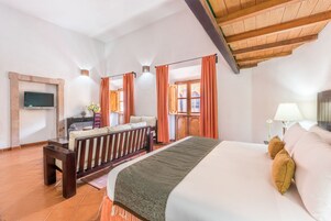 Quarto de hotel com uma cama grande, um banquinho de madeira, uma televisão e uma mesa com luminária.