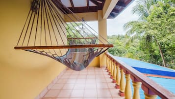 Uma rede pendurada na grade de uma varanda, com vista para uma piscina e vegetação exuberante ao fundo.