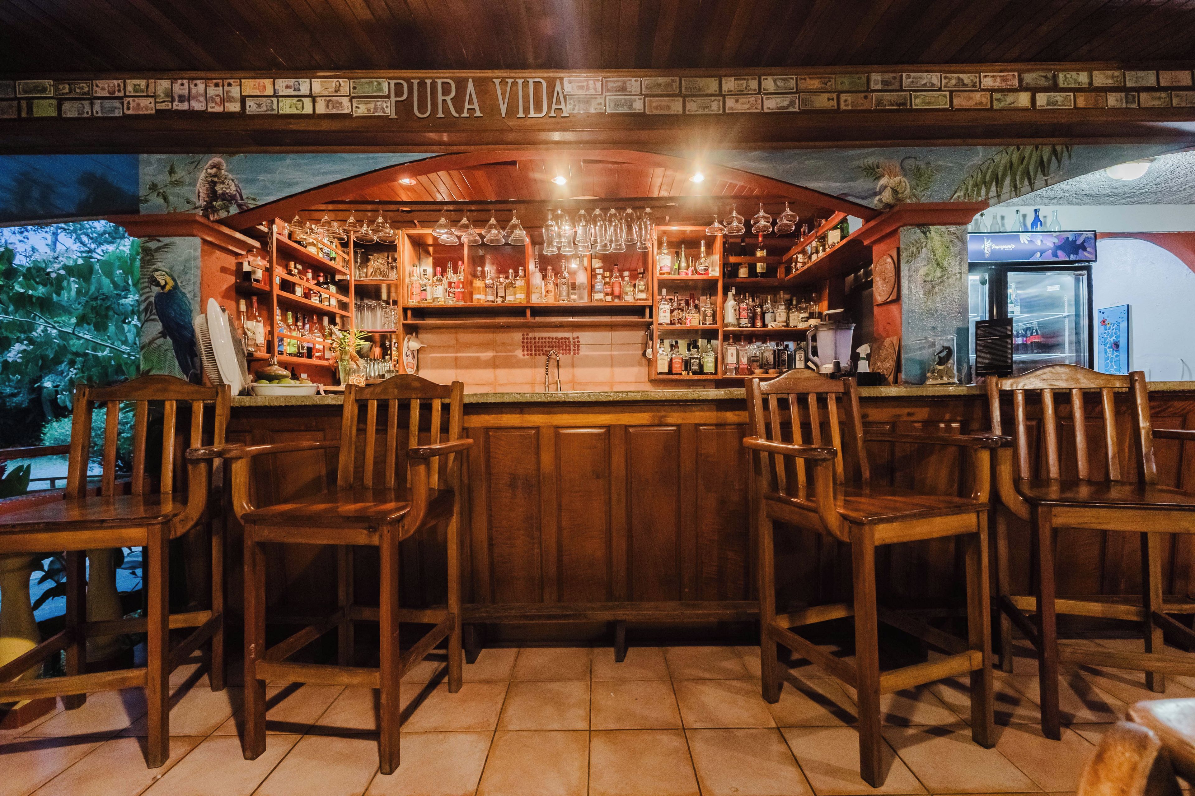 Uma área de bar com cadeiras de madeira, prateleiras cheias de garrafas e uma placa acima com a inscrição 'PURA VIDA'.