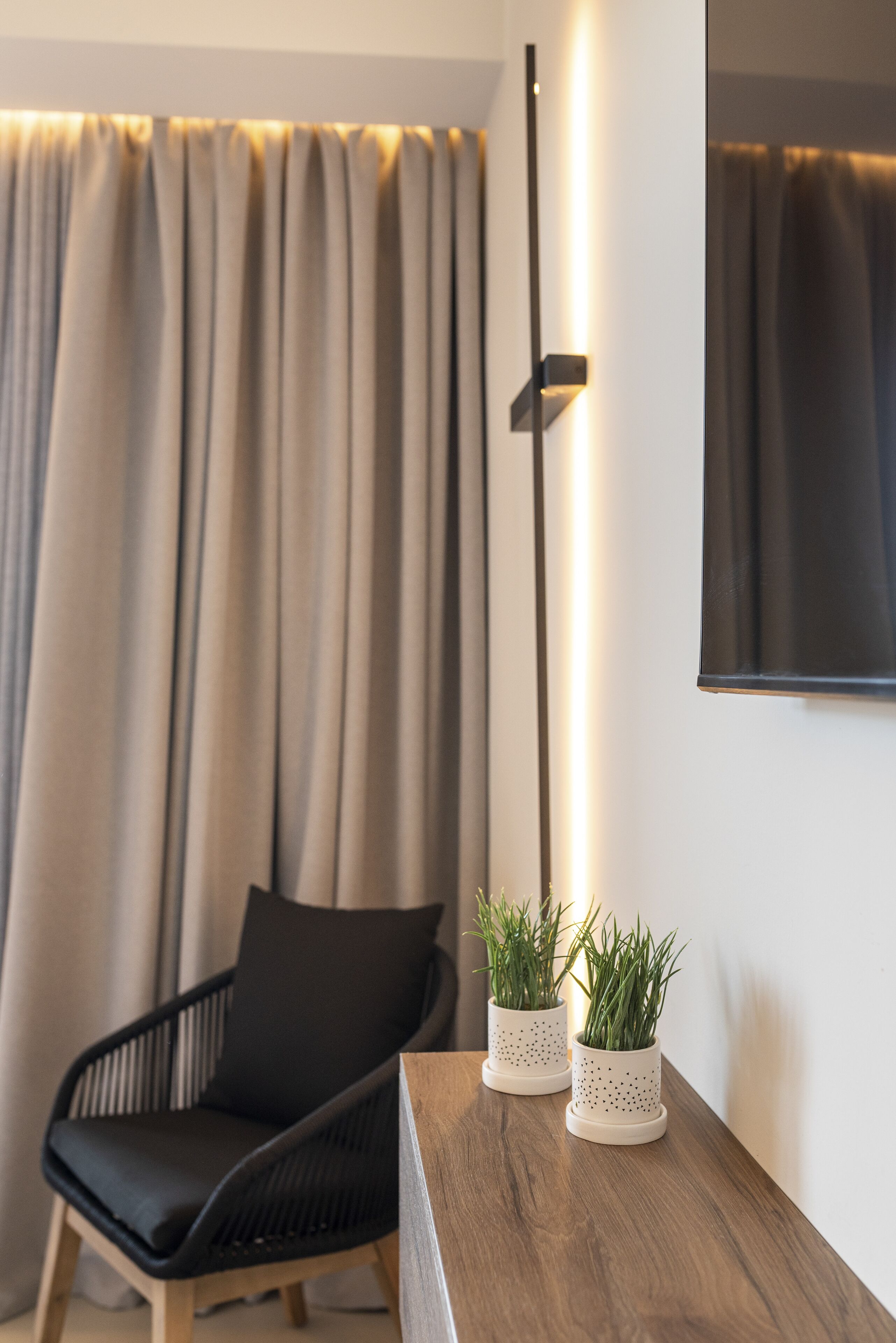 Quarto moderno com cadeira preta, duas plantas em vasos, mesa de madeira e uma janela grande com cortinas.