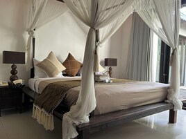 Uma cama com dossel e colunas de madeira, roupa de cama branca e almofadas decorativas. Há duas mesas de cabeceira com abajures e uma pequena mesa com um cesto e um cartão. O quarto possui uma janela grande com cortinas.