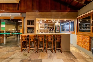 Uma área de bar com balcão de madeira, quatro banquetas e uma vitrine de vidro com garrafas.