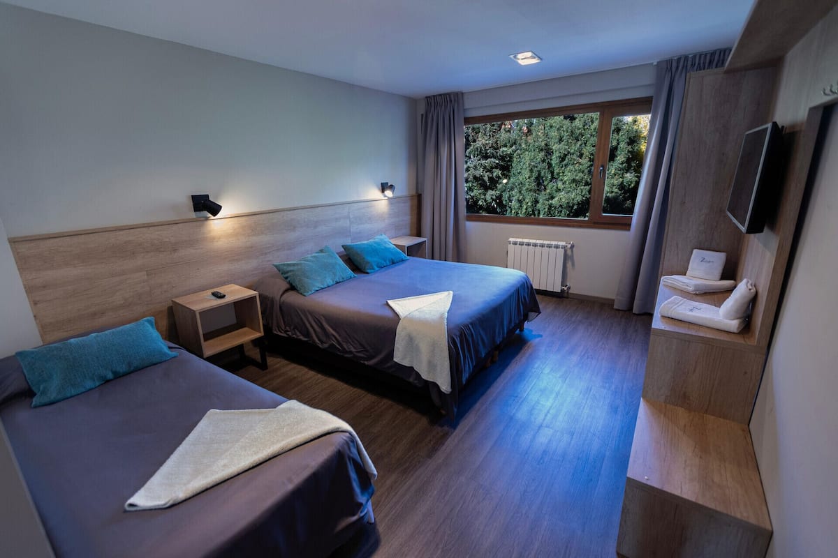 Quarto de hotel com duas camas, janela com vista para árvores, radiador e televisão fixada na parede.