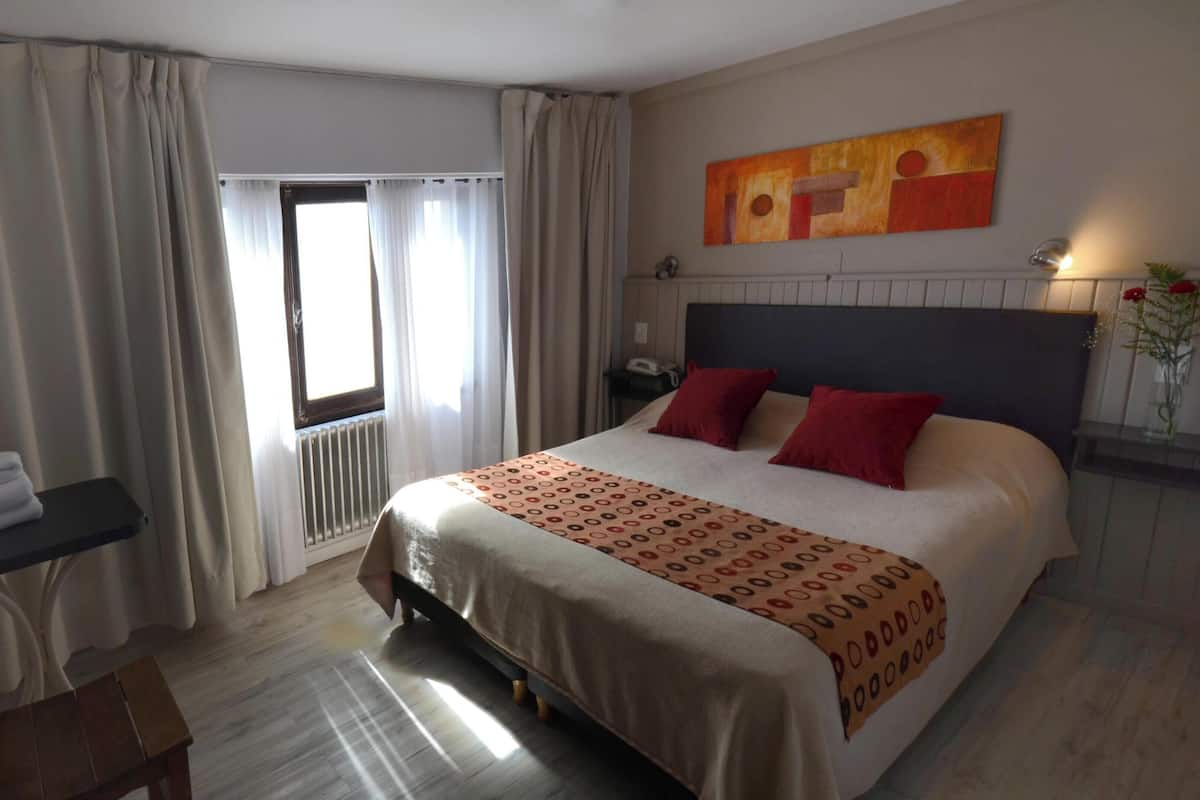 Quarto de hotel com cama, escrivaninha, cadeira, radiador e janela com cortinas.