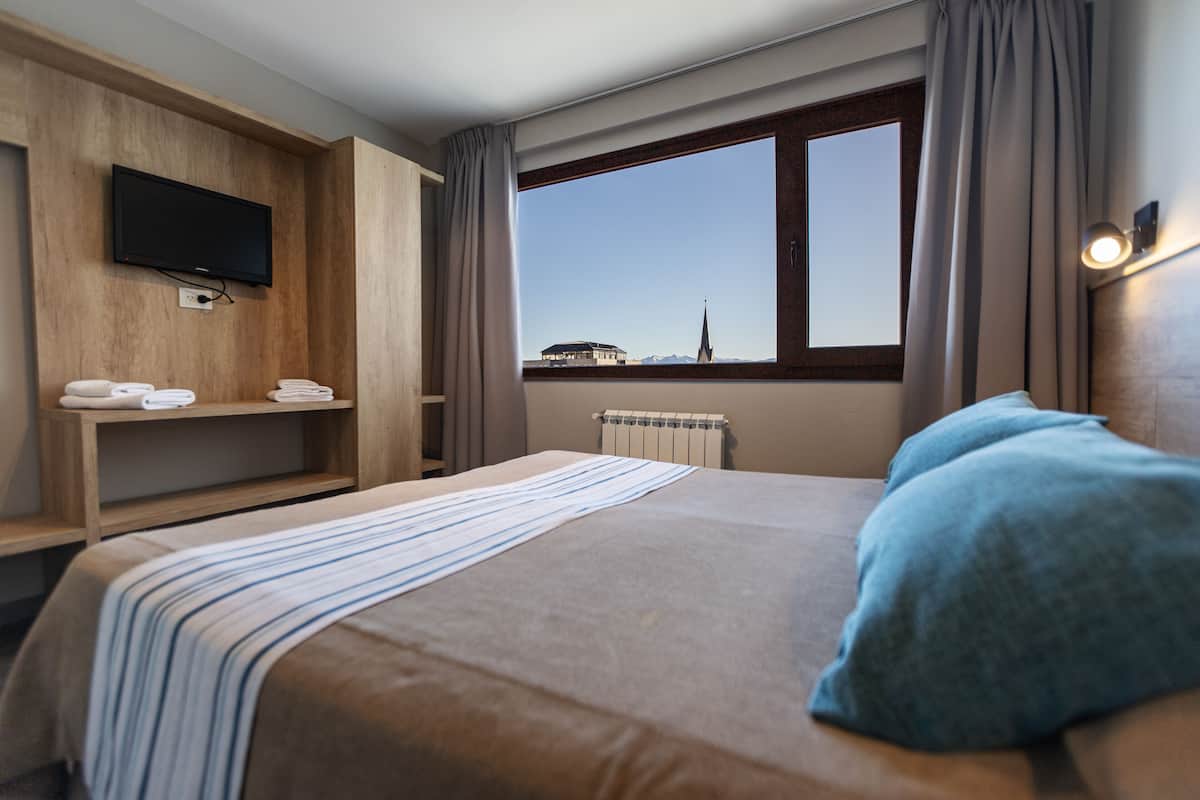 Quarto de hotel com uma cama, televisão presa à parede, janela com vista para um edifício e radiador.