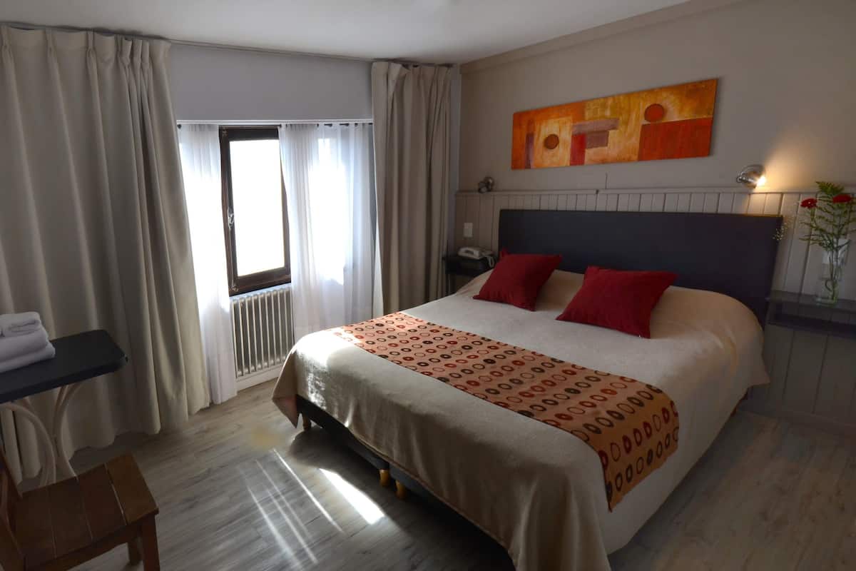 Quarto de hotel com cama, escrivaninha, cadeira, radiador e um quadro na parede.