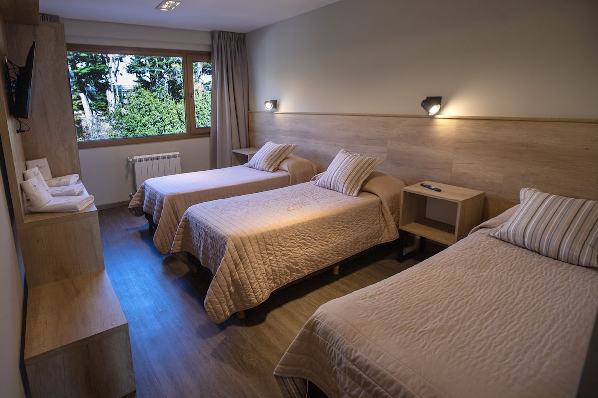 Quarto de hotel com três camas, televisão fixada na parede, janela com cortinas e mesa de cabeceira com abajur.