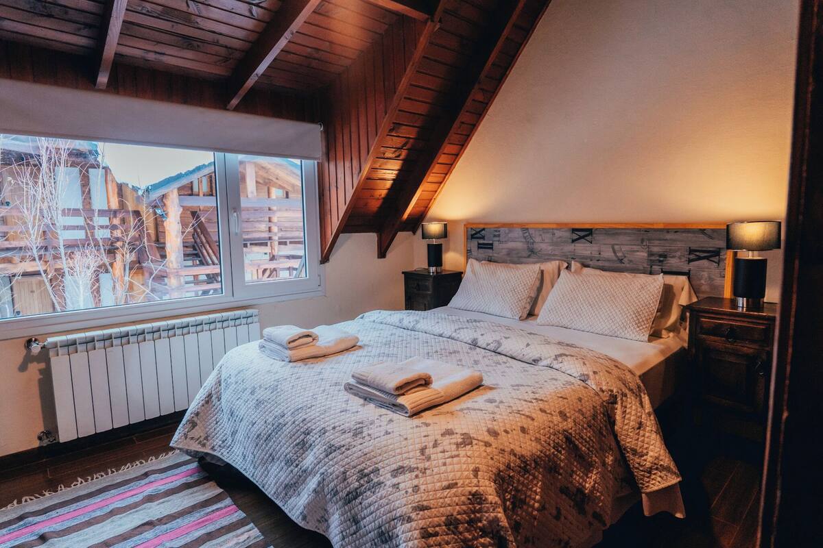 Quarto com uma cama grande, duas luminárias de cabeceira, janela com vista para edifícios e um radiador.