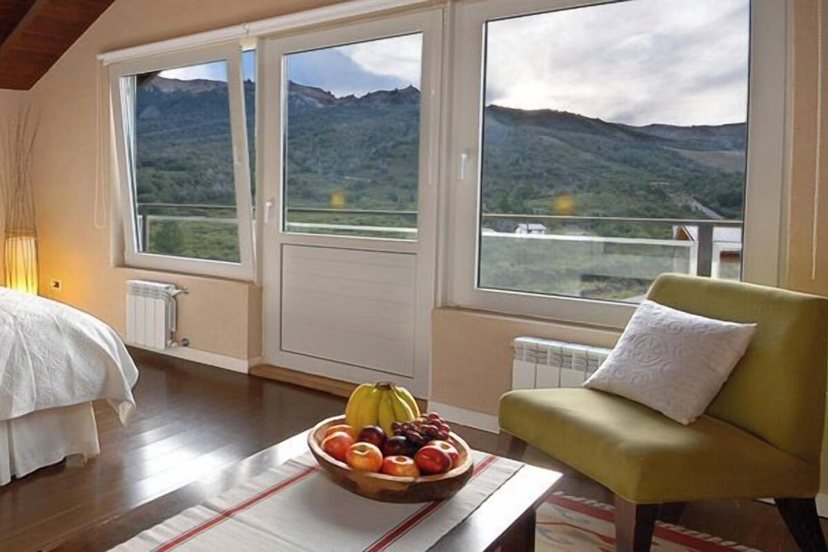 Quarto com cama, poltrona, tigela de frutas e vista para montanhas através de amplas janelas.