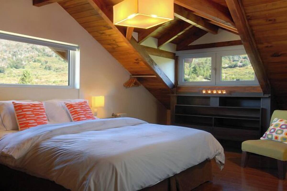 Quarto com cama, mesas de cabeceira, uma cadeira e duas janelas.