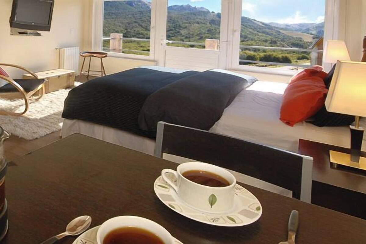 Quarto aconchegante com cama, cafeteira, xícara de café e vista para montanhas.