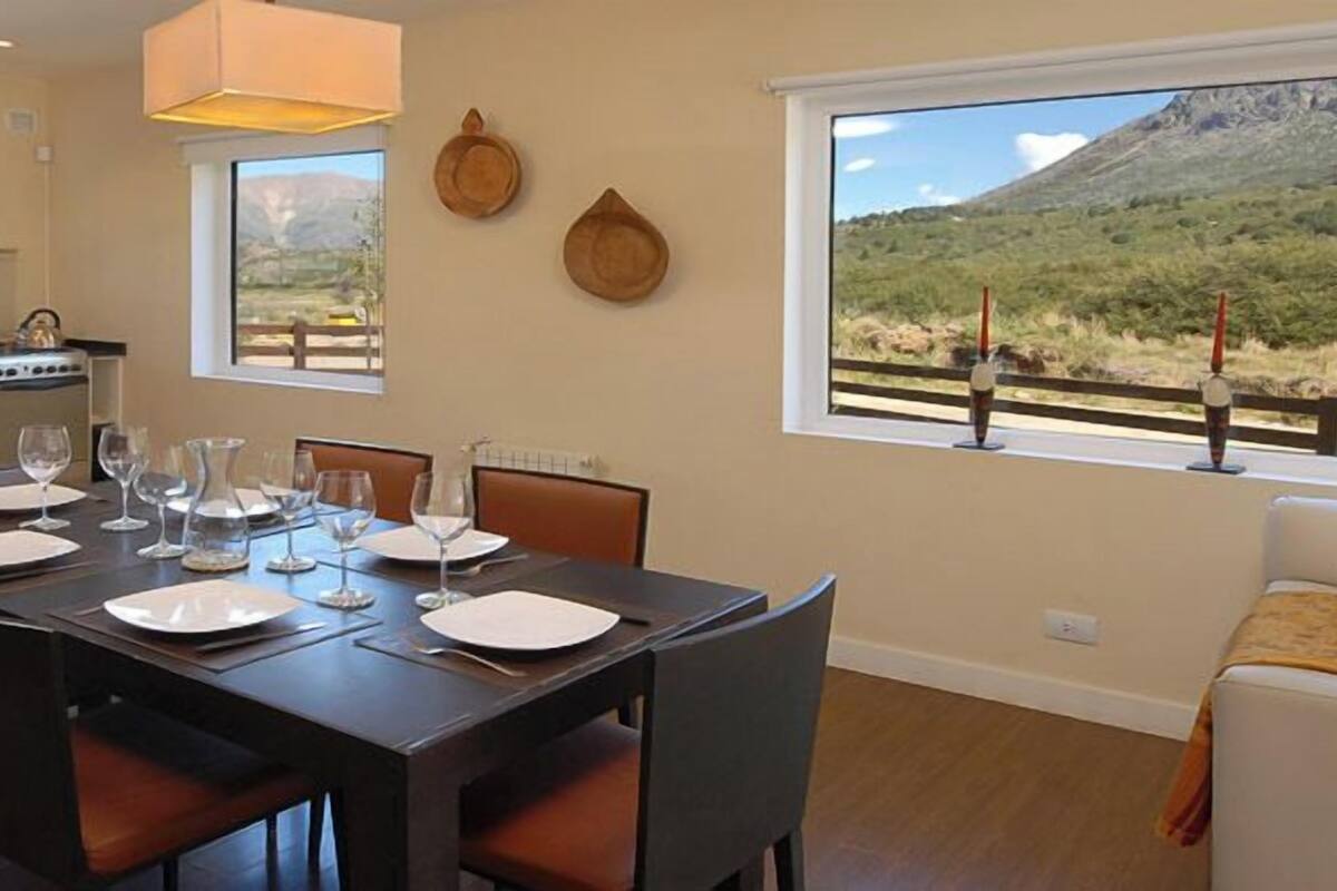Uma sala de jantar com mesa posta para quatro, uma cozinha com armários brancos e uma janela grande com vista para as montanhas.