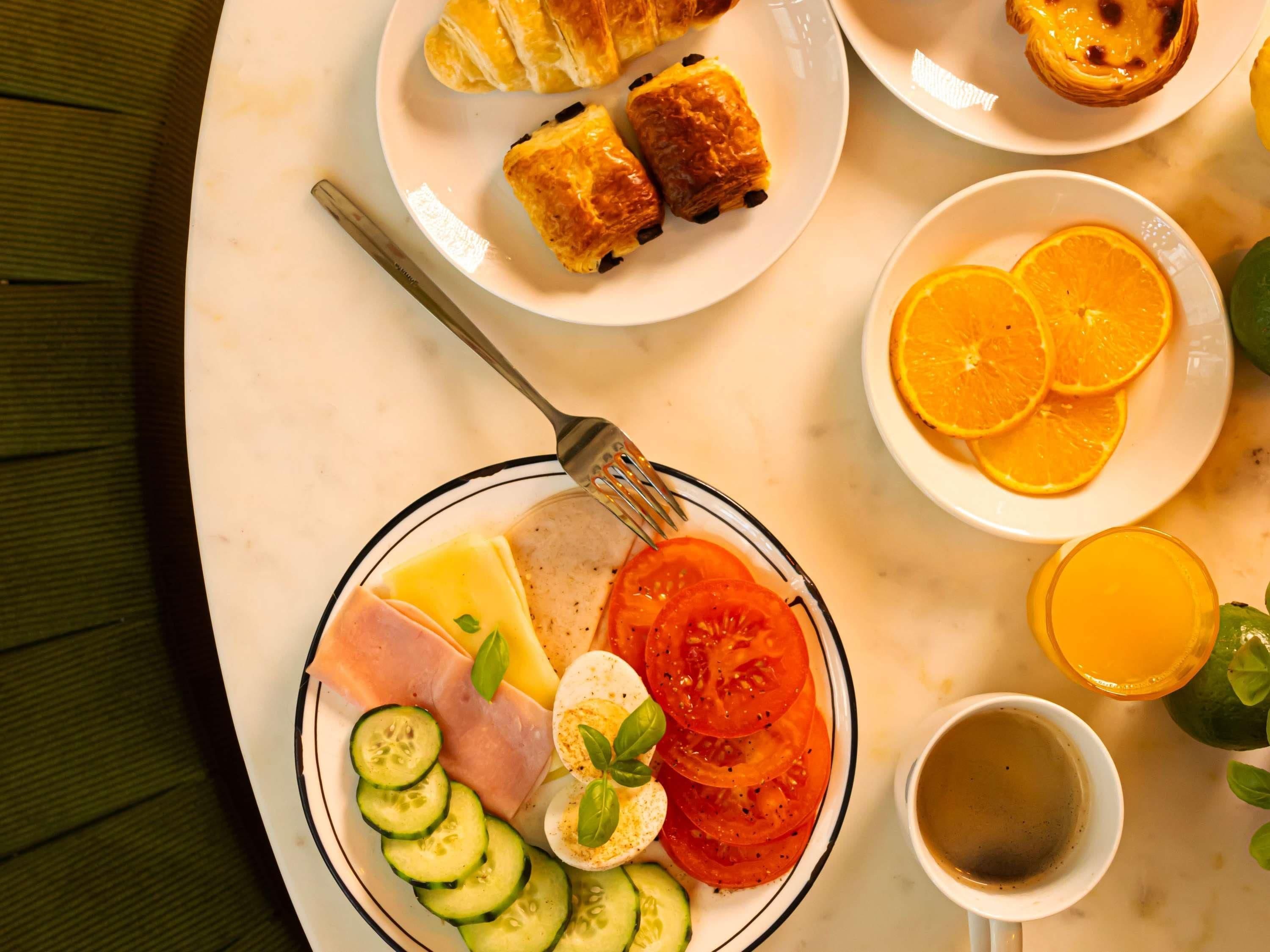 Mesa de café da manhã com um prato de tomates, pepinos, queijo e presunto, um prato de fatias de laranja, um copo de suco de laranja e uma xícara de café.
