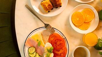 Mesa de café da manhã com um prato de tomates, pepinos, queijo e presunto, um prato de fatias de laranja, um copo de suco de laranja e uma xícara de café.