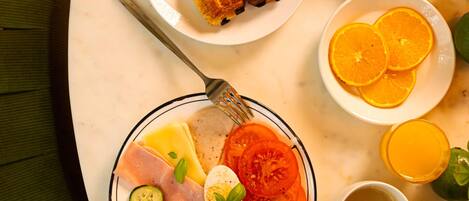 Mesa de café da manhã com um prato de tomates, pepinos, queijo e presunto, um prato de fatias de laranja, um copo de suco de laranja e uma xícara de café.