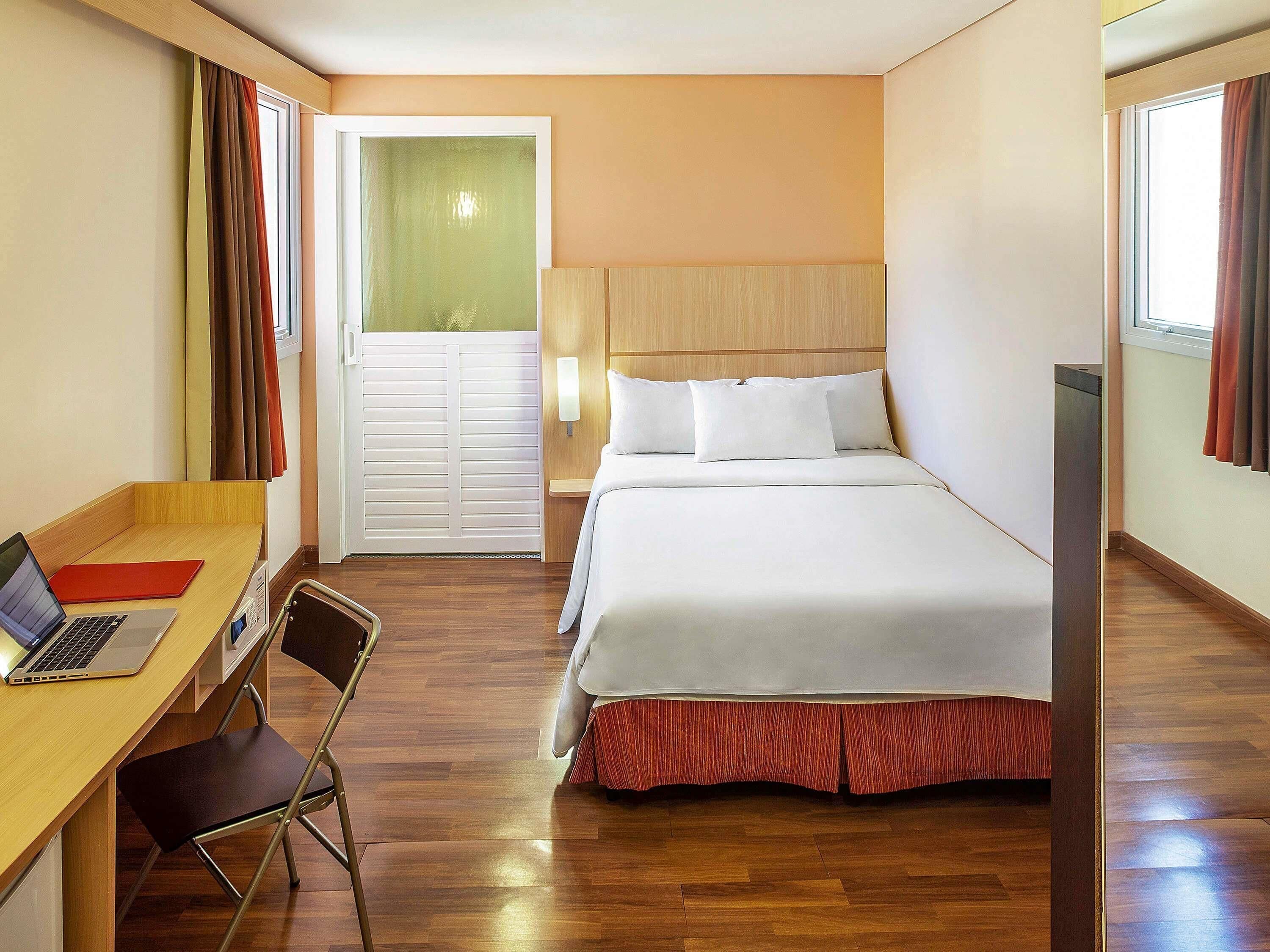 Quarto de hotel com cama, escrivaninha com um laptop, cadeira e porta para uma varanda.
