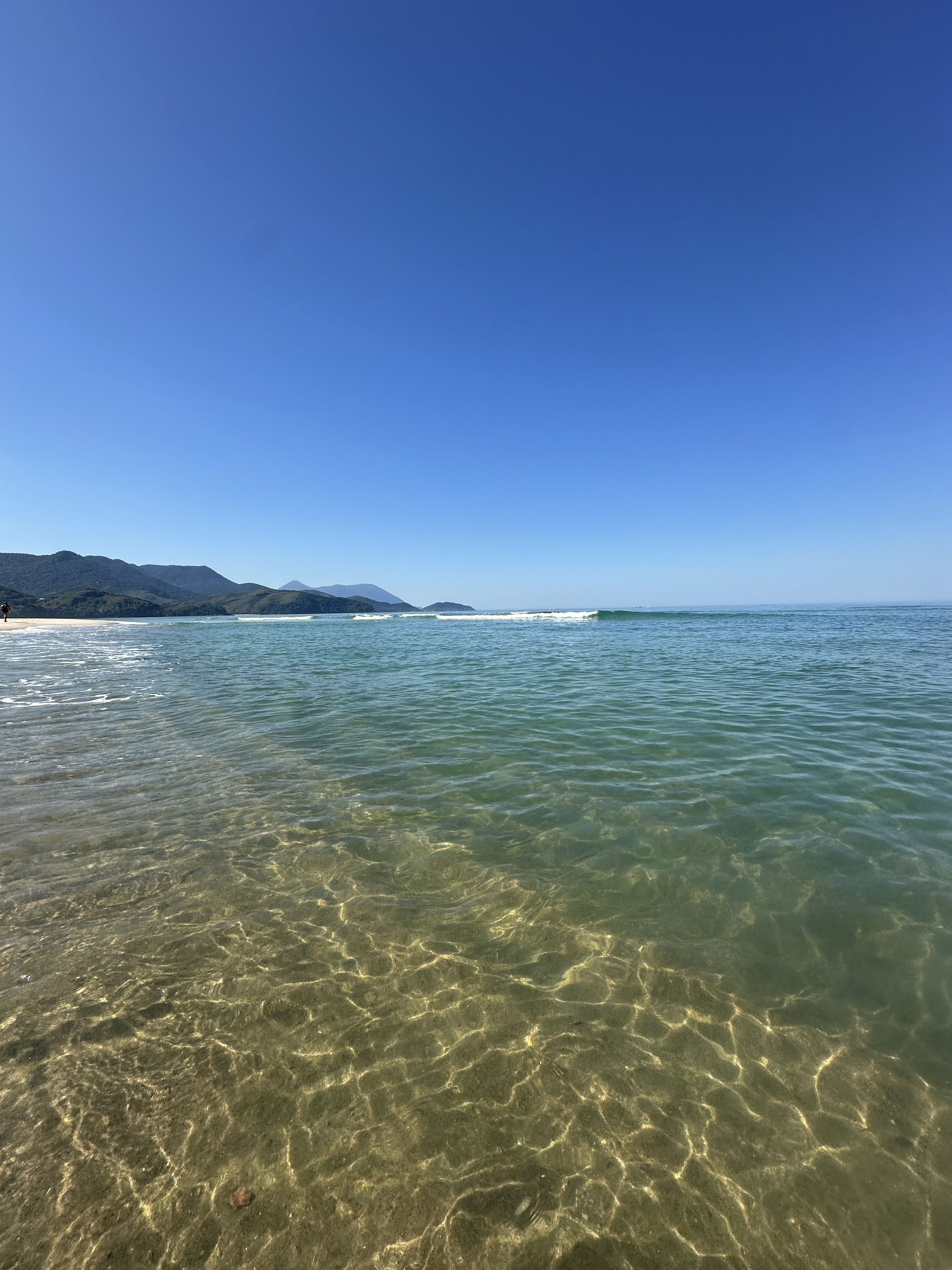 Vista clara de uma praia com ondas suaves e uma cordilheira ao longe.