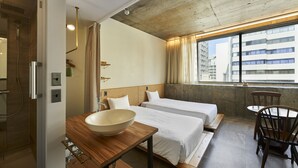 Quarto de hotel com mesa de madeira, pia com bacia, duas camas, uma cadeira e vista da cidade através de grandes janelas.