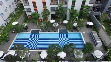 Piscina no terraço com padrão quadriculado, cercada por árvores e guarda-sóis, em frente a um edifício moderno com varandas.