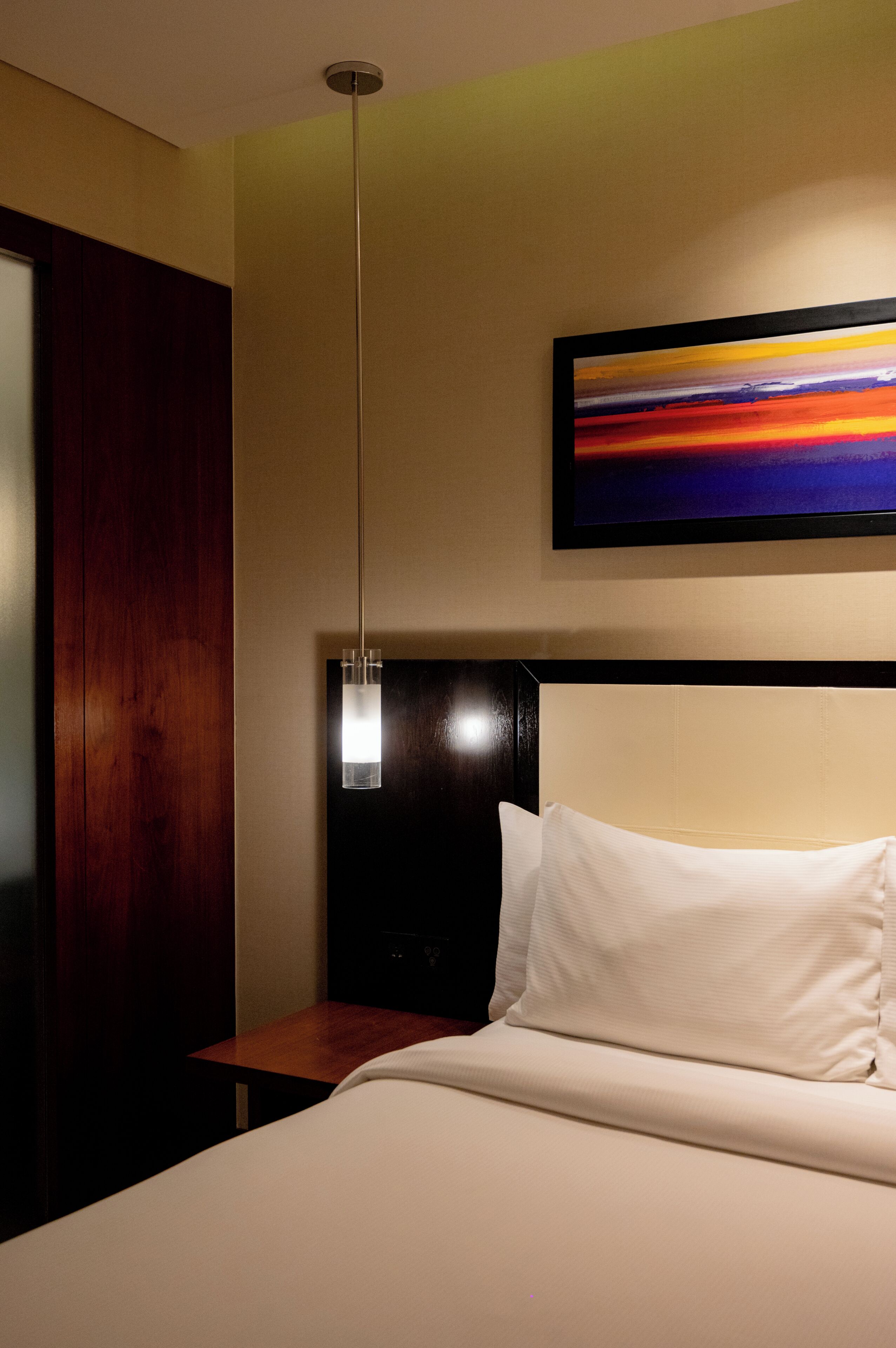 Quarto de hotel com cama, mesa de cabeceira, um quadro e uma luminária pendente.