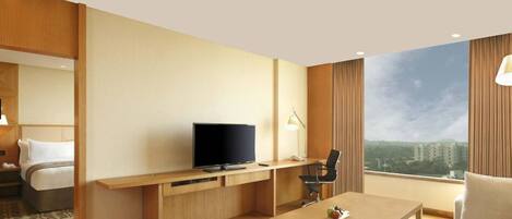 Quarto de hotel moderno com cama, TV em suporte de madeira, escrivaninha, cadeira e vista para a cidade através de uma janela grande.