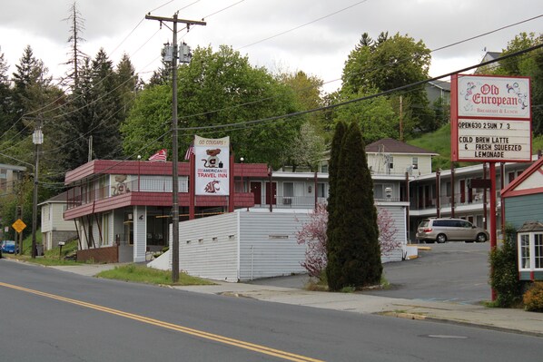 Um motel com fachada vermelha e branca, um letreiro anunciando 'Old European Breakfast & Lunch', e um estacionamento com um carro.