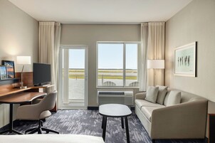 Quarto de hotel com escrivaninha, cadeira, sofá e vista do exterior através de amplas janelas.