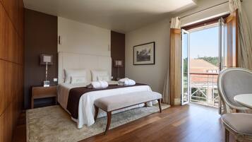 Quarto de hotel com cama, mesas de cabeceira, um banco, uma cadeira e vista para o exterior.