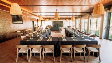 Sala de conferências com mesas compridas, cadeiras e luminárias pendentes.
