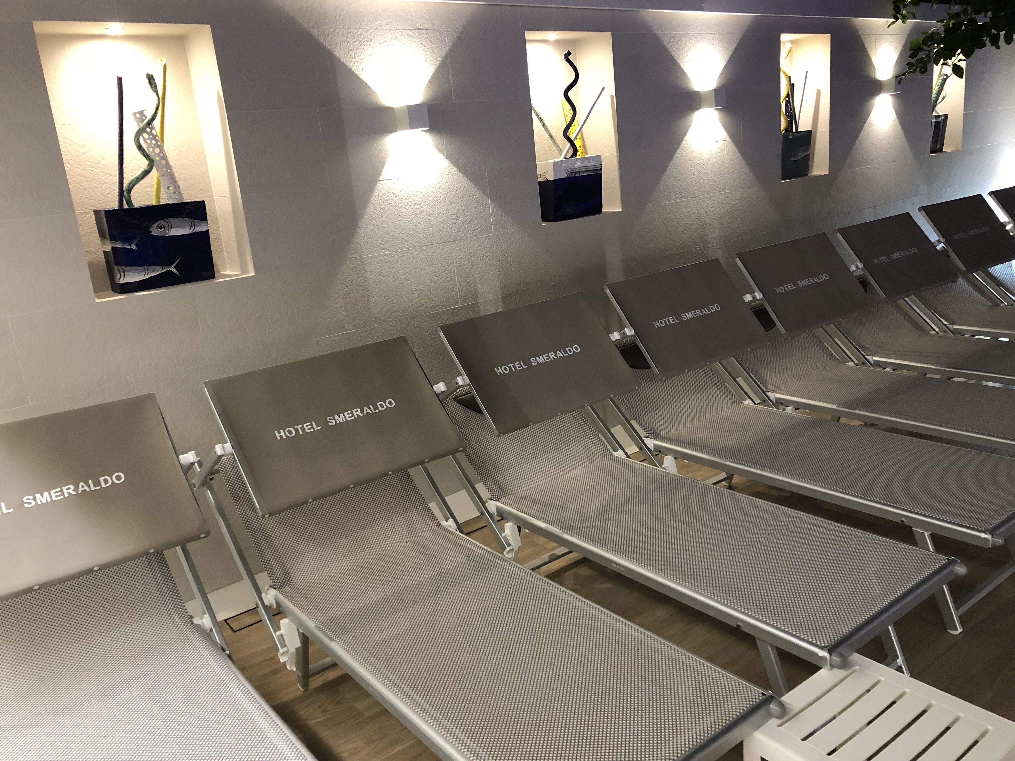 Um saguão de hotel com fileiras de poltronas de descanso, identificadas como "HOTEL SMERALDO", e luminárias fixadas na parede.