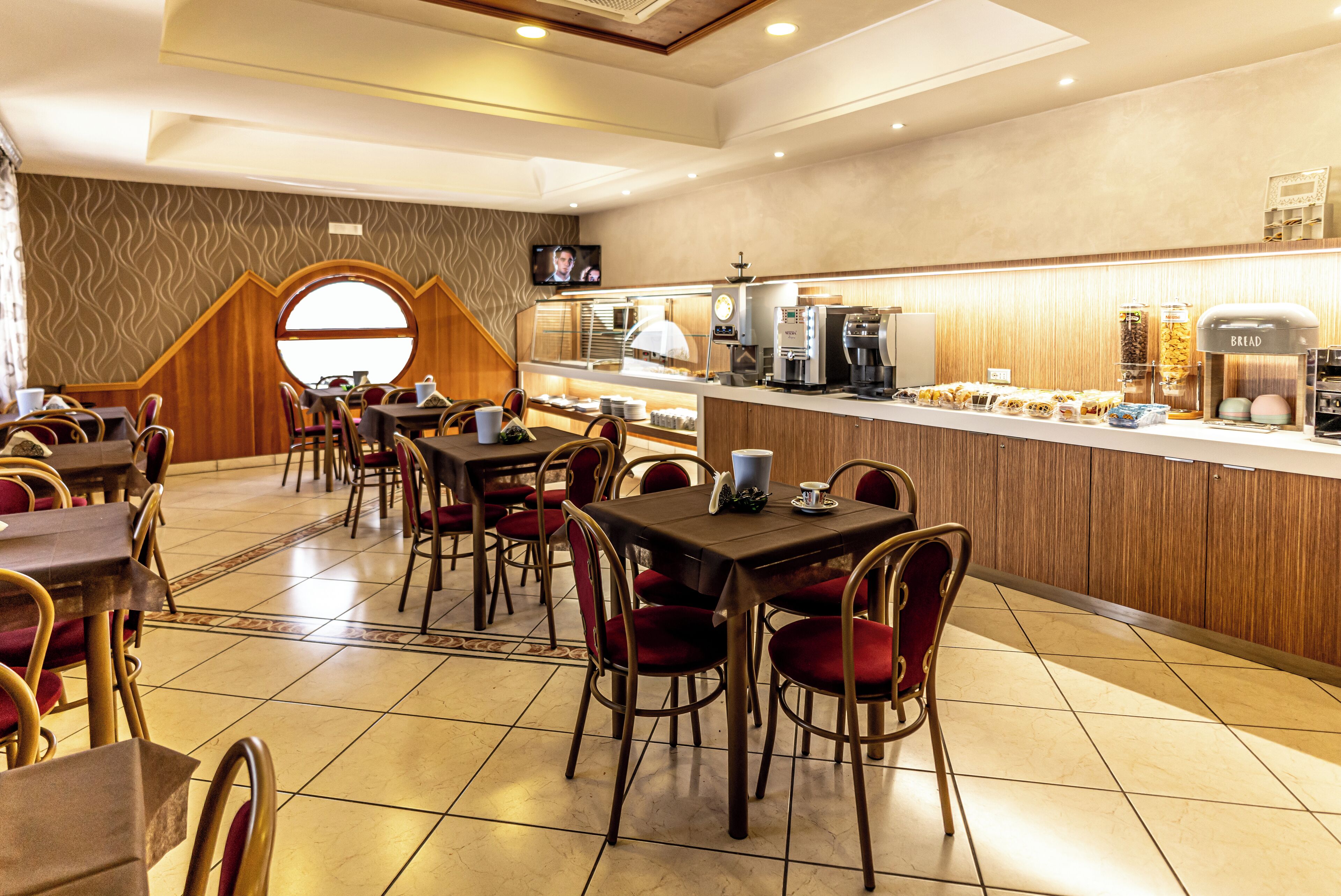 Uma sala de jantar de hotel com mesas, cadeiras, um canto de café e uma tela de televisão.