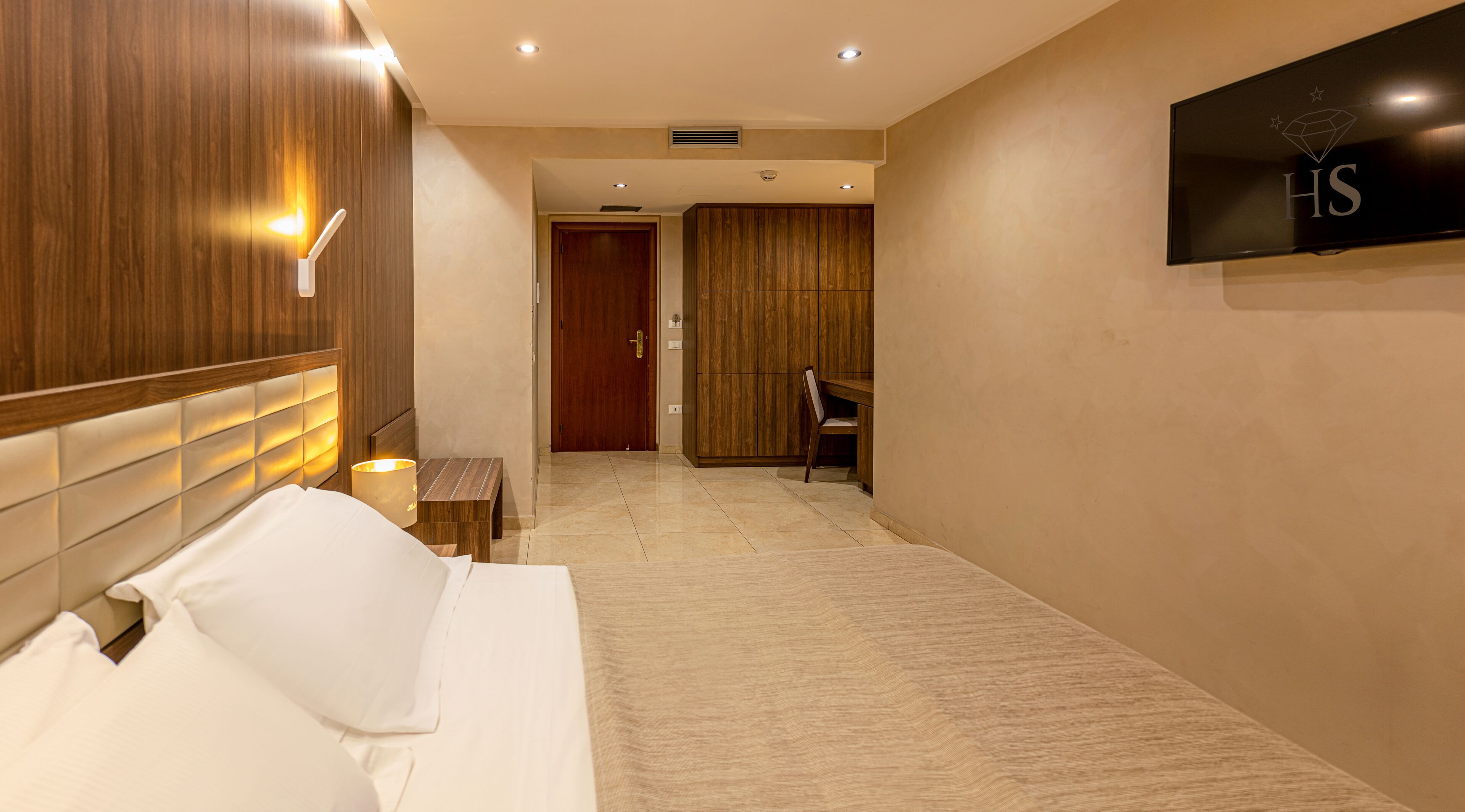 Quarto de hotel com cama, mesa de cabeceira, luminária, guarda-roupa de madeira e televisão presa na parede.