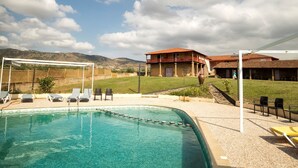 Piscina cristalina com espreguiçadeiras e área de estar coberta, rodeada por edifícios e colinas.