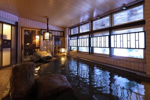 Uma onsen japonesa tradicional com interior de madeira, uma nascente quente de pedra e grandes janelas que permitem a entrada de luz natural.